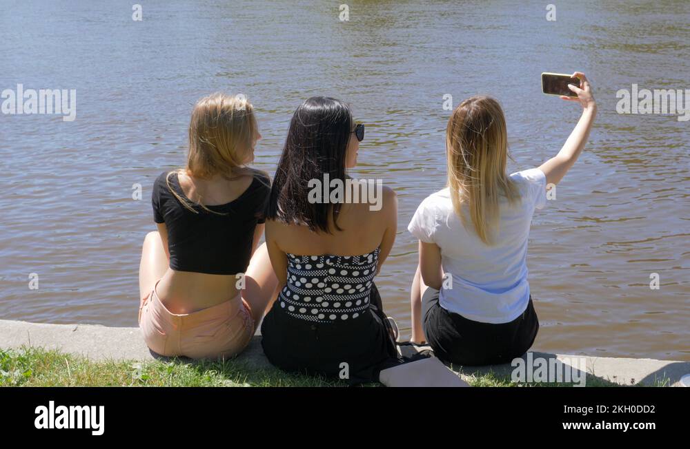 Three young girls friends back view sit together near river water ...