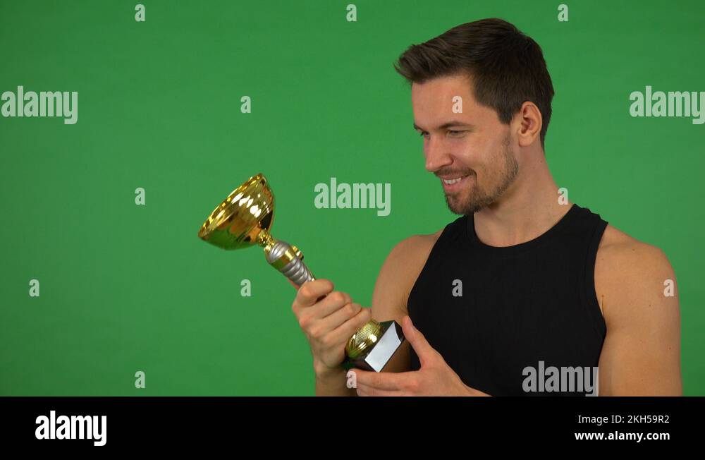 A young handsome athlete holds a golden trophy and celebrates - green ...