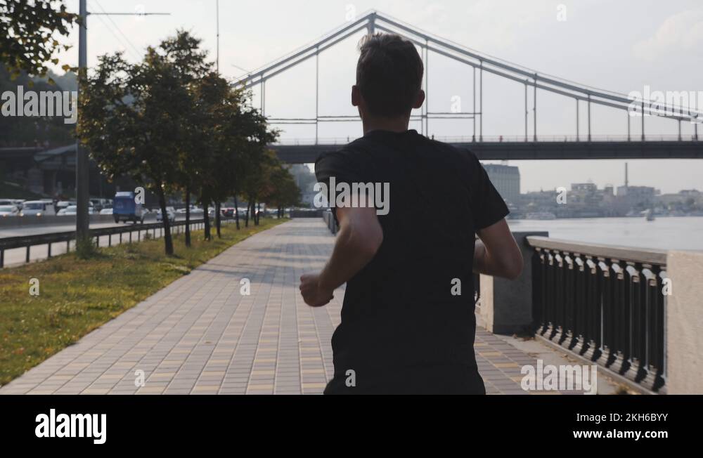 Back side view of young slim man running along river on city quay ...