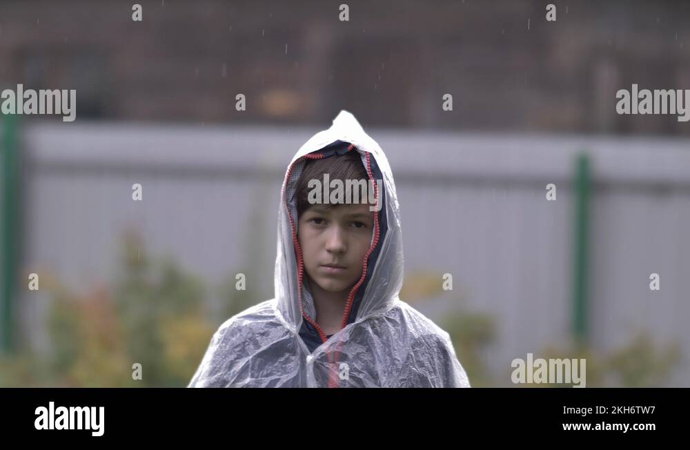 boy with a sad face is standing in a raincoat in the rain, pain on face ...