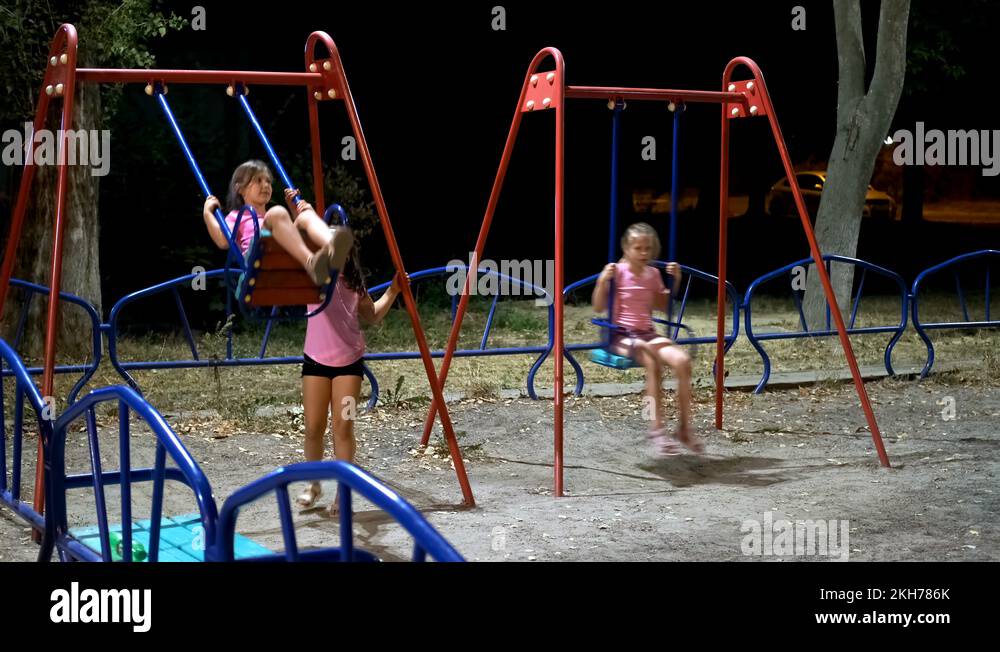 Three girls at playground enjoying at night in open air. Girls swing ...