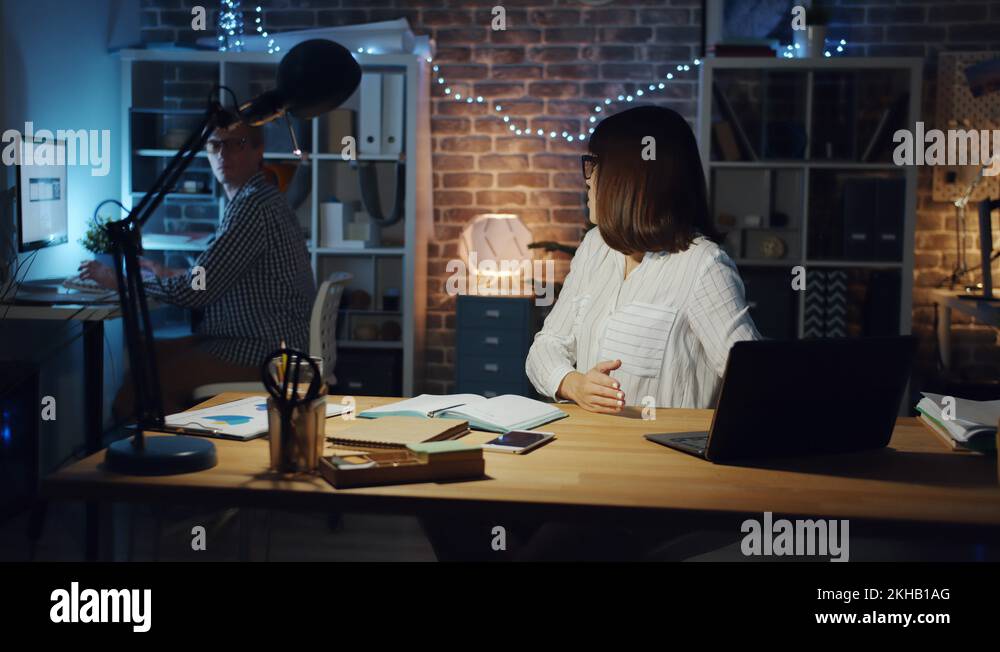 Joyful man helping female colleague with computer work talking in dark ...