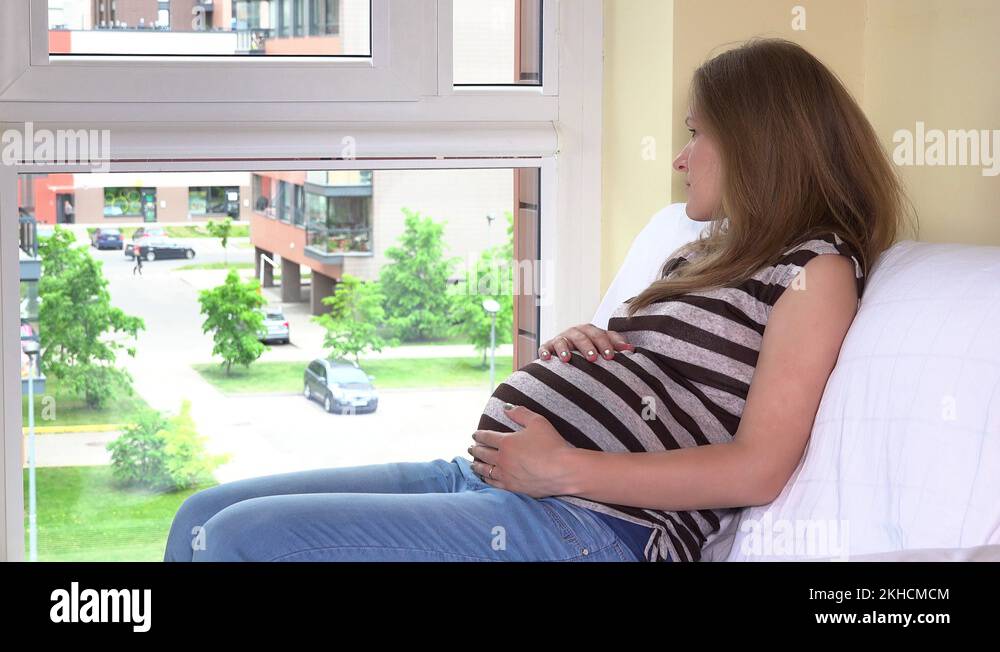 Young pregnant woman caress tender her stomach and look through window ...