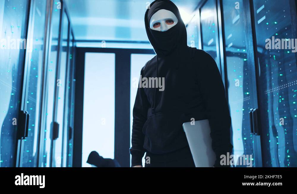 Hooded anonymous hacker with laptop standing inside server room ...