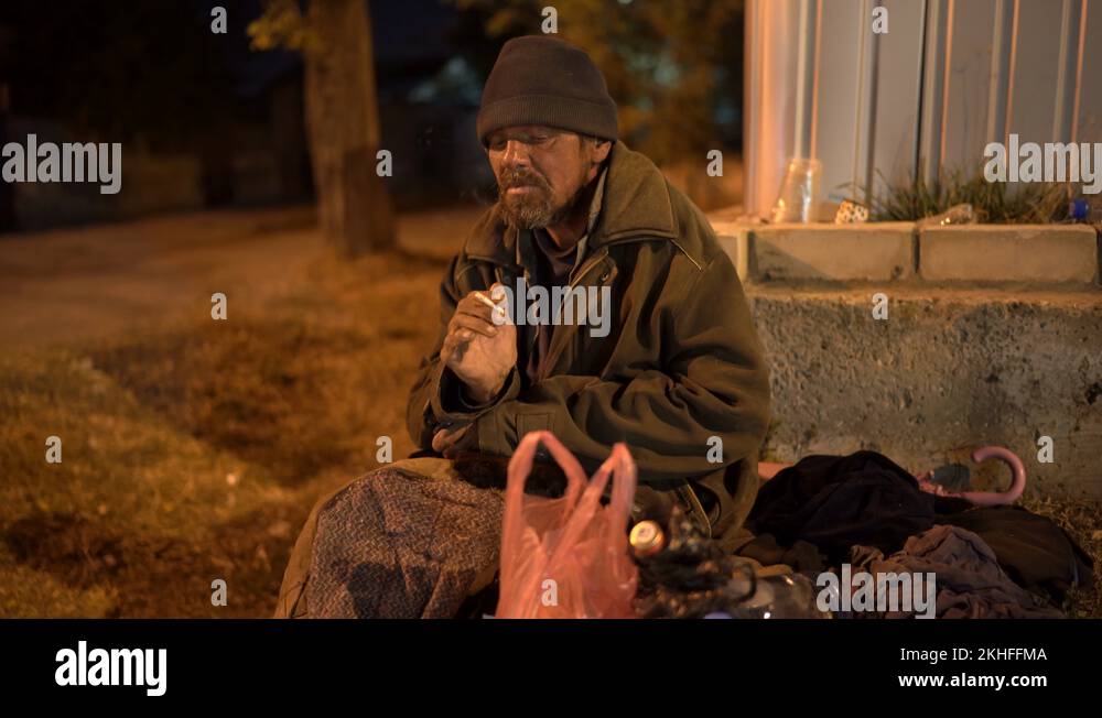 Lonely cold homeless man sitting on the street at night smoking ...