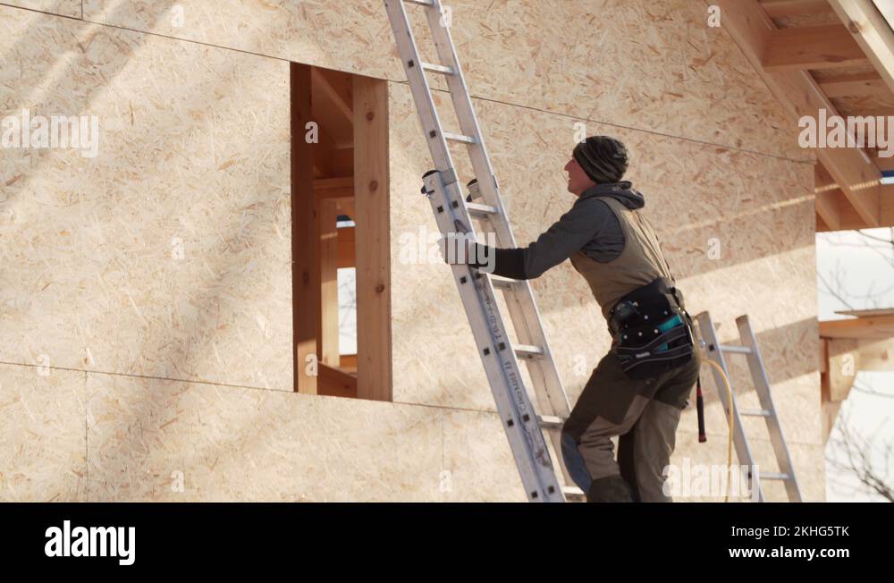 Builder climbs high ladder with hydraulic hammer. Frame house under ...