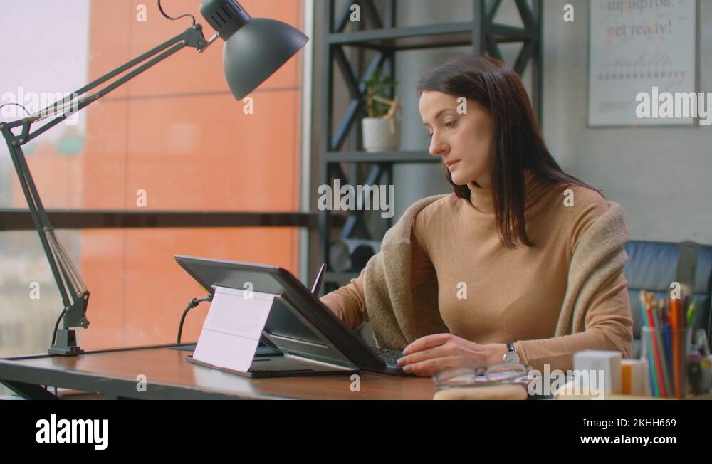 A female graphic designer works in an office with large Windows and ...