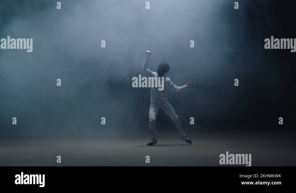 Professional fencer in uniform practicing maneuvers with sword in dark ...