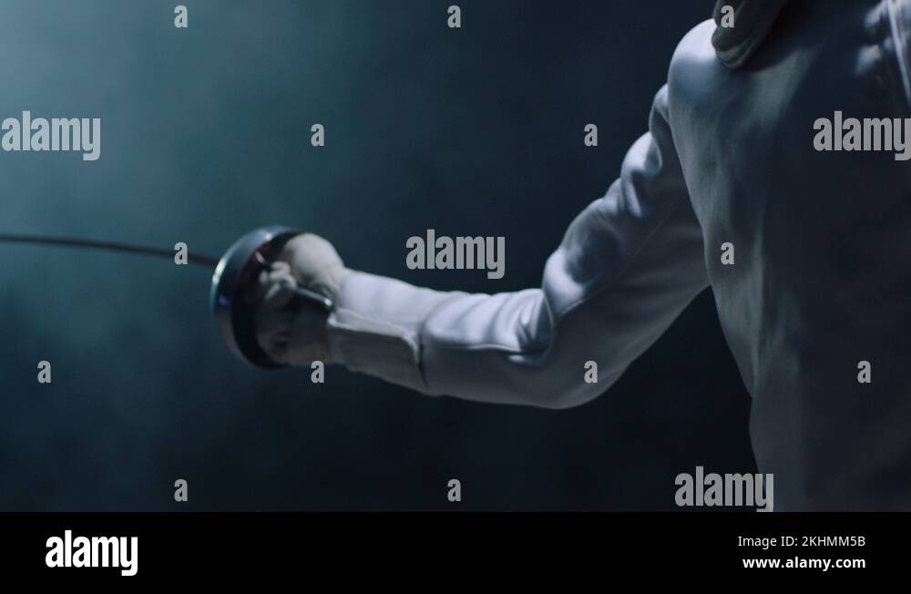 Professional fencer in uniform practicing maneuvers with sword in dark ...