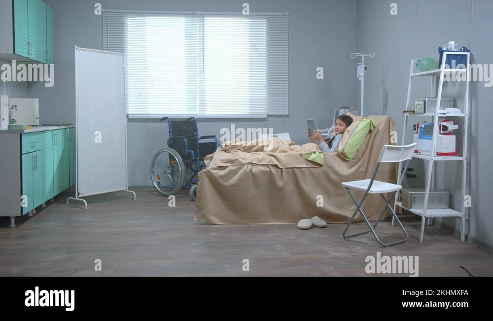 Girl lie on the bed in the hospital, hold a tablet in hands and look ...