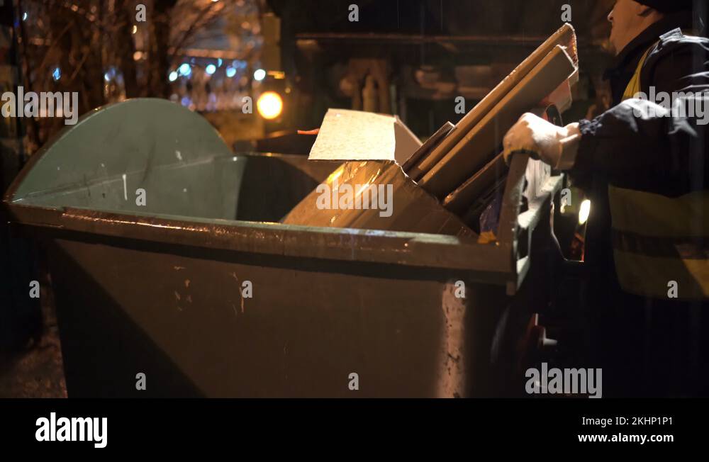 Garbage man at work on rainy city street at night, garbage truck lifts ...
