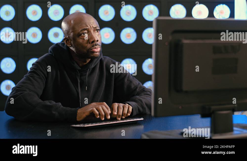 Bald bearded african-american male hacker in black hoodie types on ...