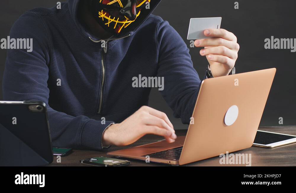 Male hacker in a hood uses laptop to access credit card in dark office ...