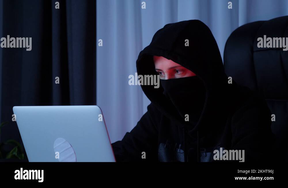 Female face in black mask and hoodie looking at laptop screen. Hacker ...