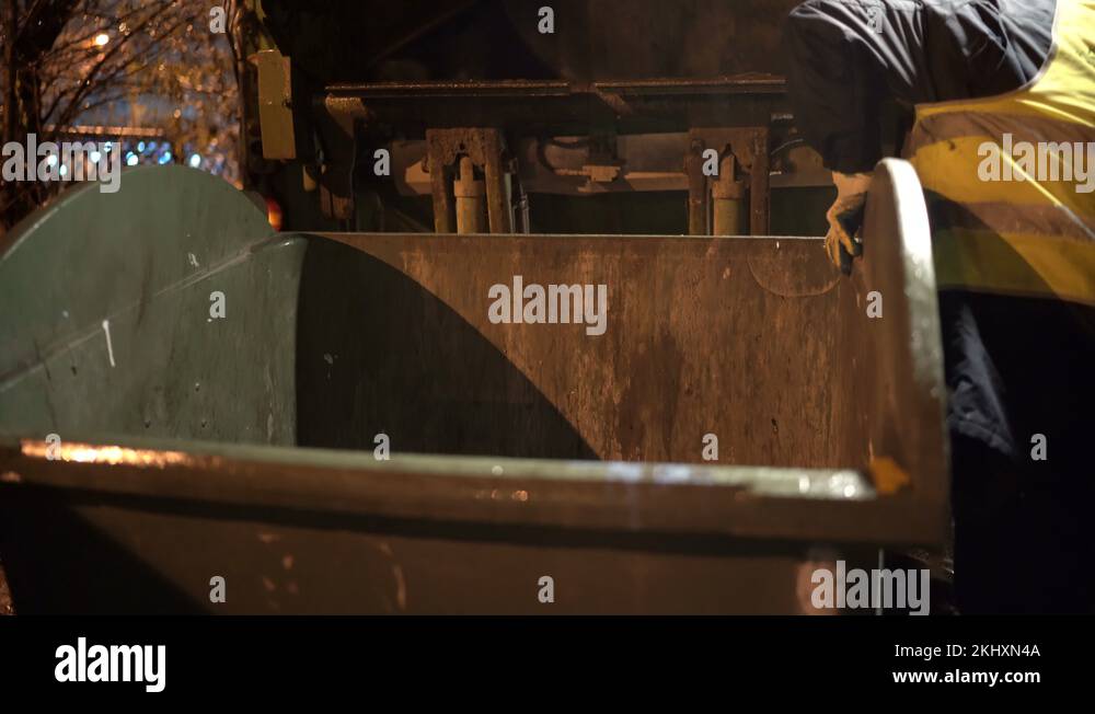 Garbage man at work on rainy city street at night, garbage truck lifts ...