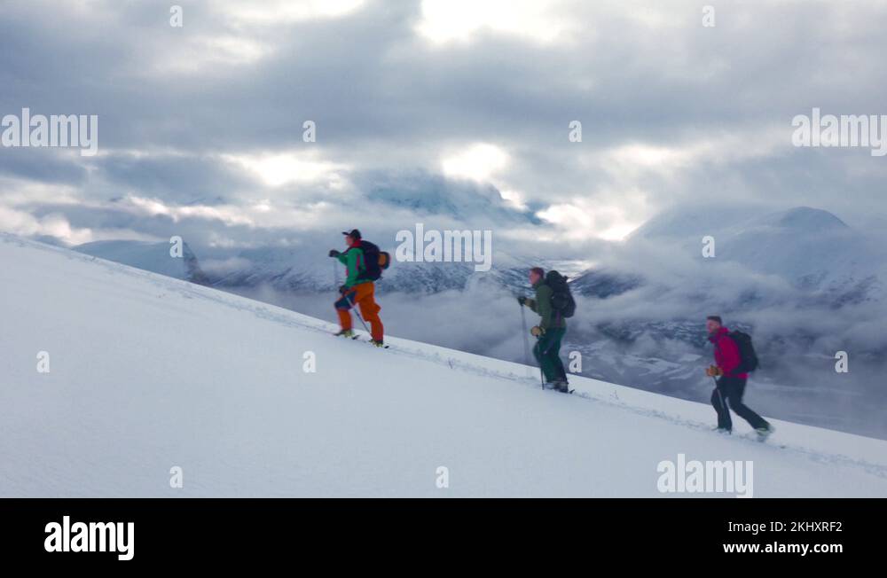 Three men walking uphill, wearing ski's, in the snow on a huge mountain ...