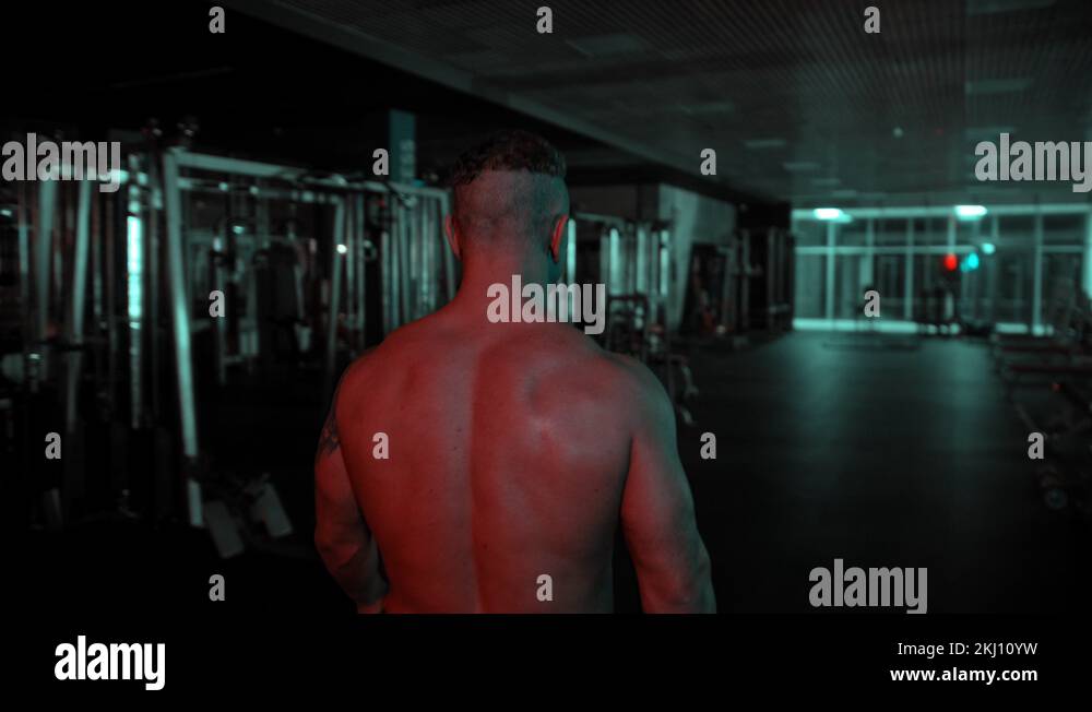 An adult shirtless man bodybuilder walking in the dark gym with neon ...
