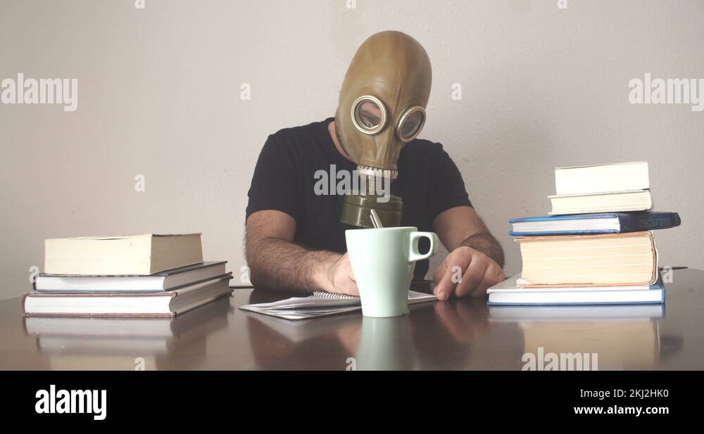 Man in a gas mask writing in a notebook between books at an office ...