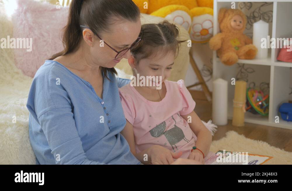 A woman helps her daughter learn to read a book, sitting together on ...