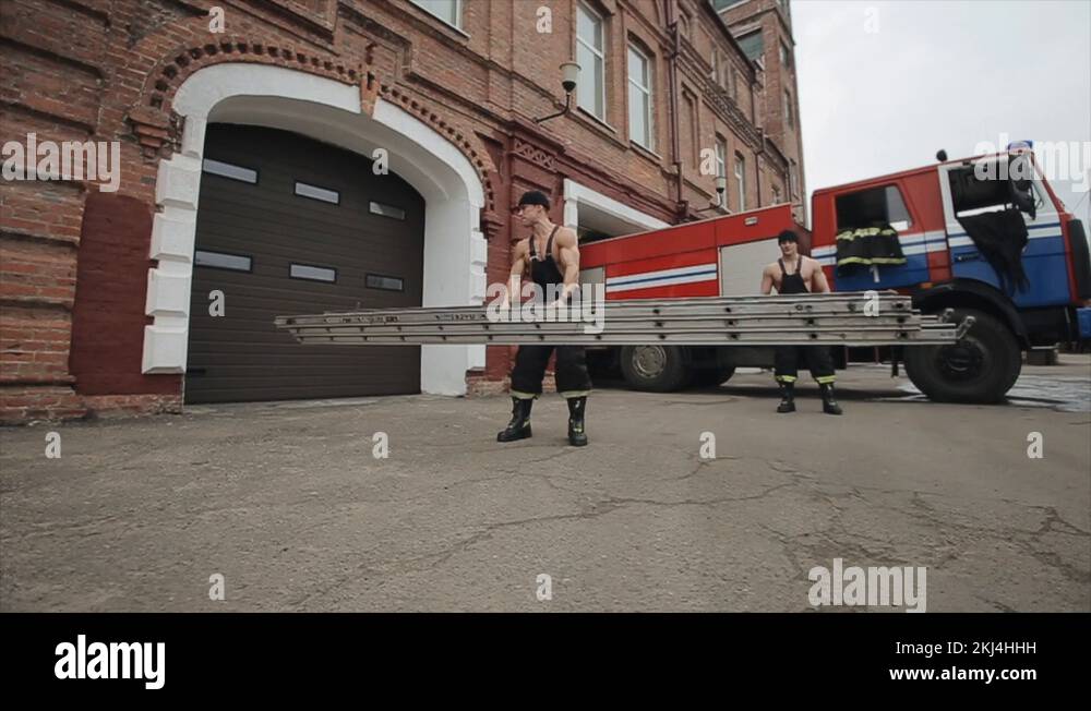 Two muscular young fireman train with the help of a long metal ladder ...