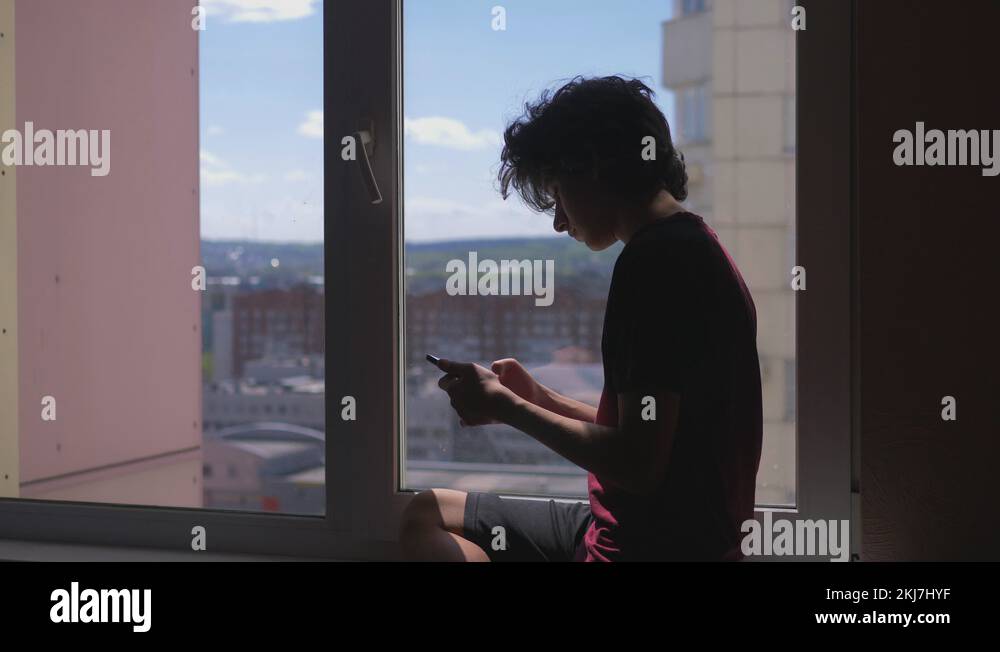 silhouette. teenage boy uses smartphone sitting on window sill in his ...