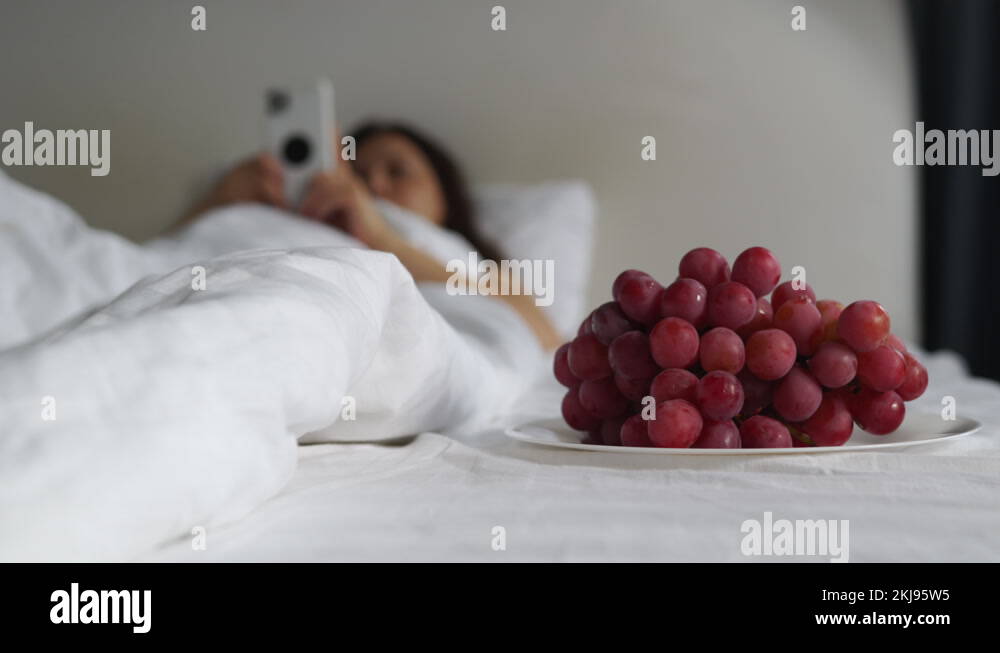Relaxed woman lying in bed using mobile phone and eating fresh grapes ...