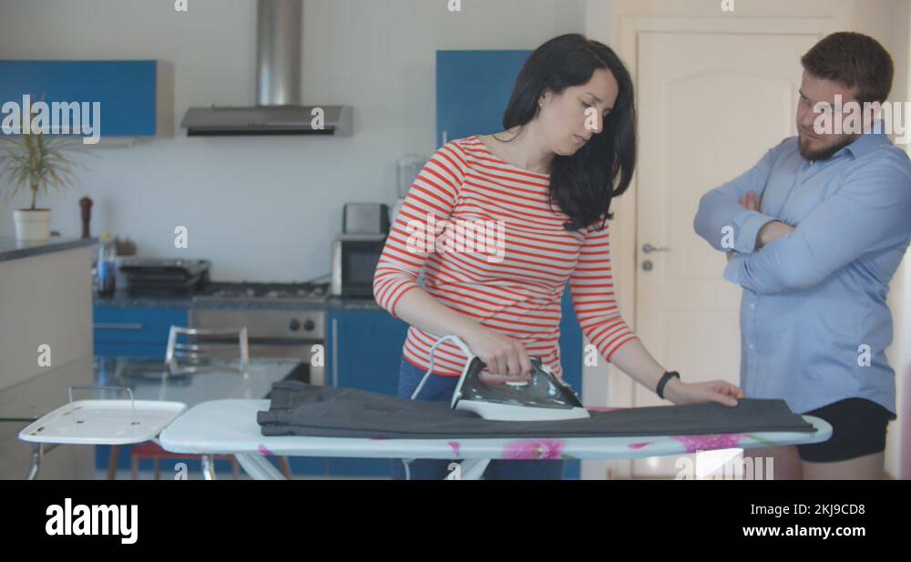 Angry man in shorts and a shirt waiting while his wife ironing trousers ...