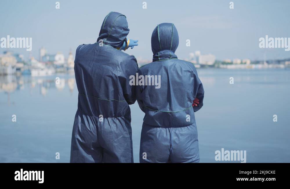 Back view of two people in biohazard suits standing at riverbank and ...