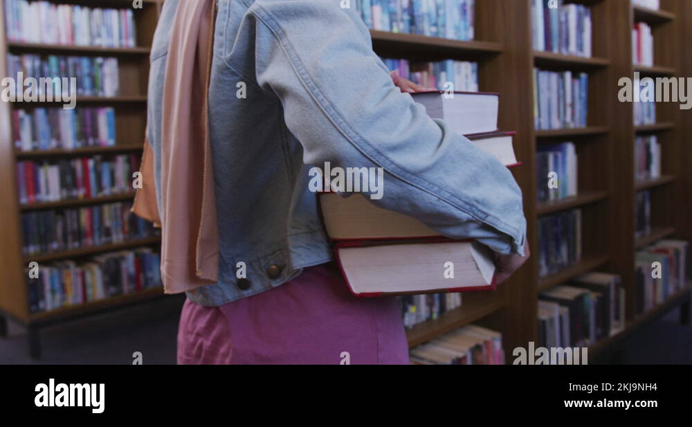 Asian female student walking between bookshelves and carrying a stack ...