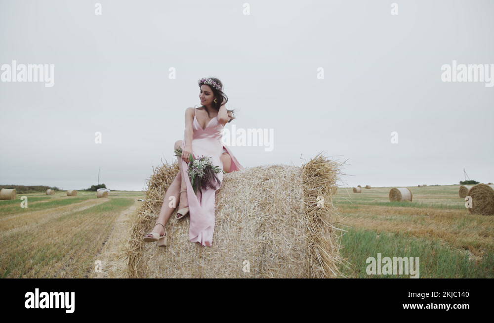 Happy elegant lady sits on a large haystack in a windy field and smiles ...
