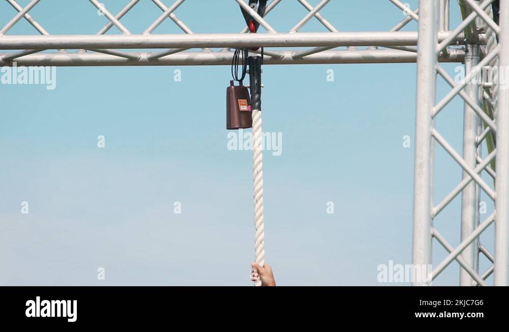 Woman climbing tall rope and reaching up to hit bell during obstacle ...