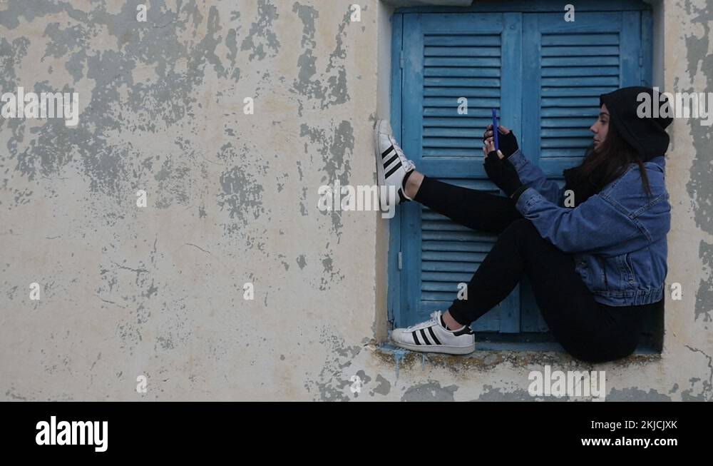 A teenage girl sits on the outside of a shuttered window while using ...