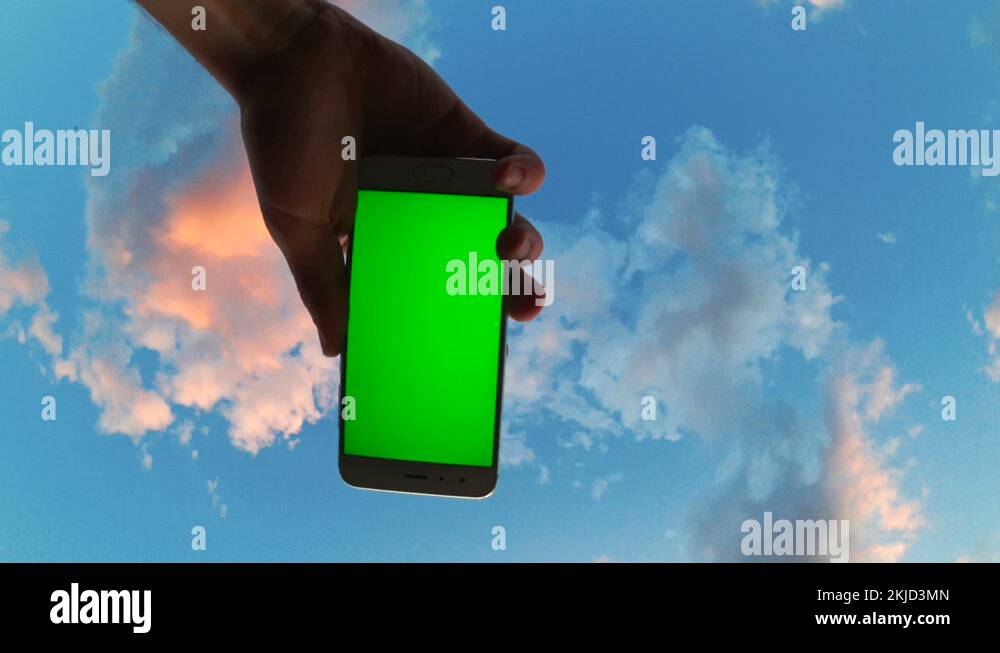 Man's hand holding mobile smartphone with green screen on cloudy sky ...