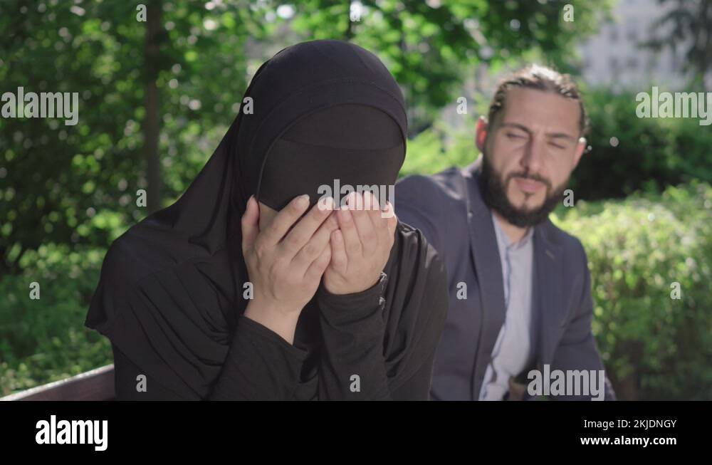 Close-up of crying muslim woman in hijab sitting on bench as blurred ...