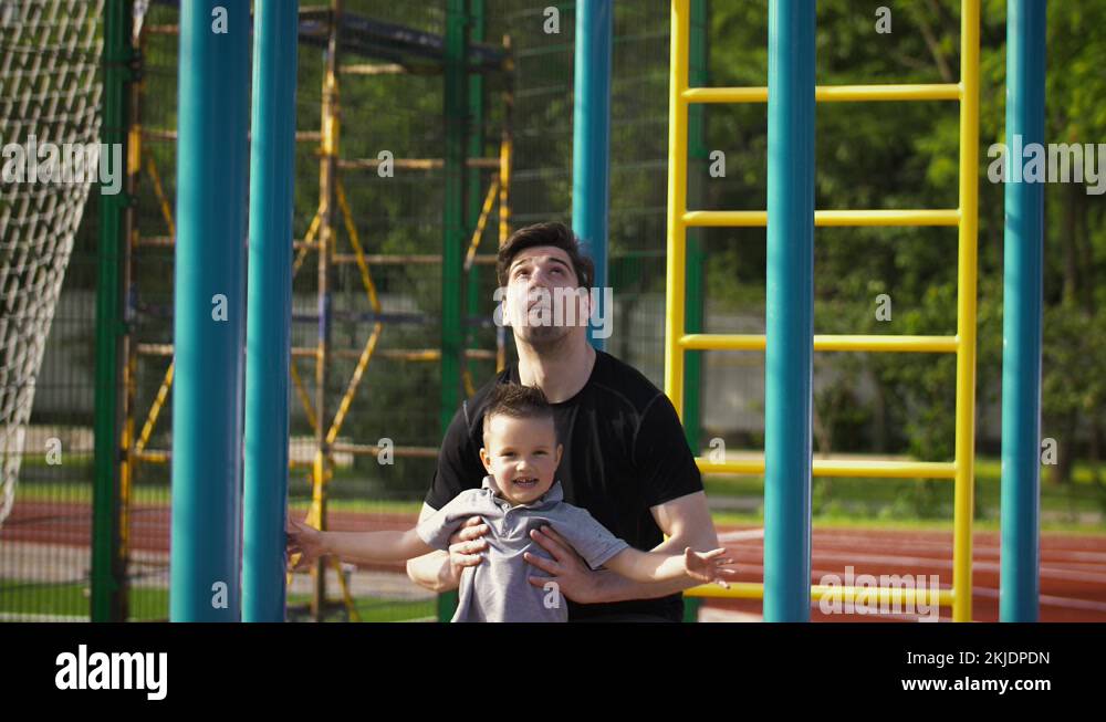 Dad in sportswear raising child boy on crossbar of the sports complex ...