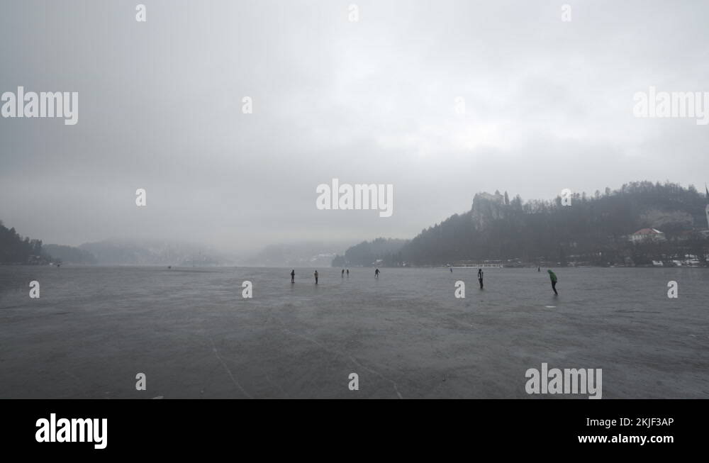 Pan of people walking and skating on frozen Lake Bled below Castle and ...