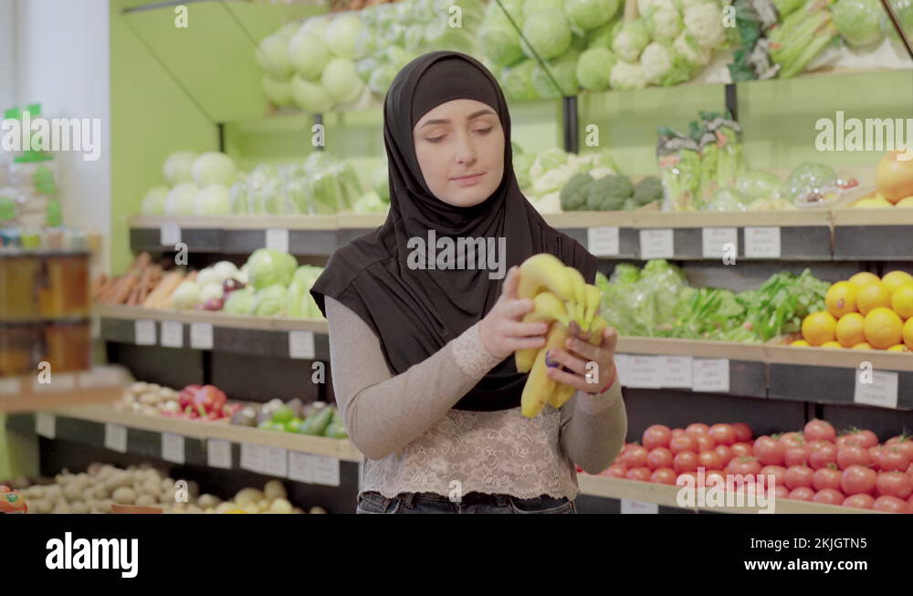 Young Muslim woman examining bunch of bananas in grocery. Portrait of ...