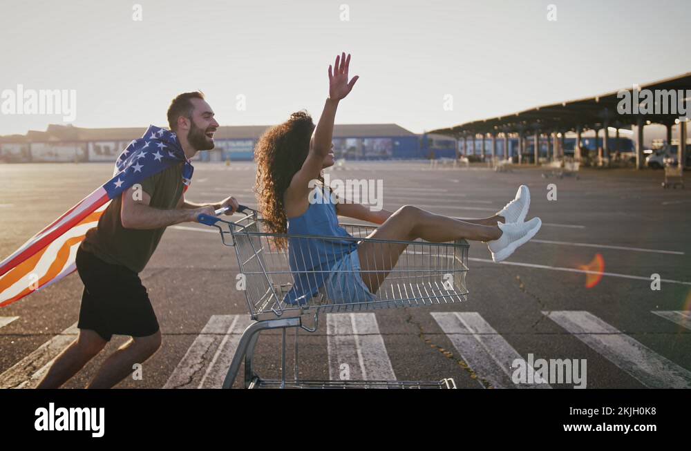 Young guy wrapped in flag of USA is racing on shopping trolley with ...