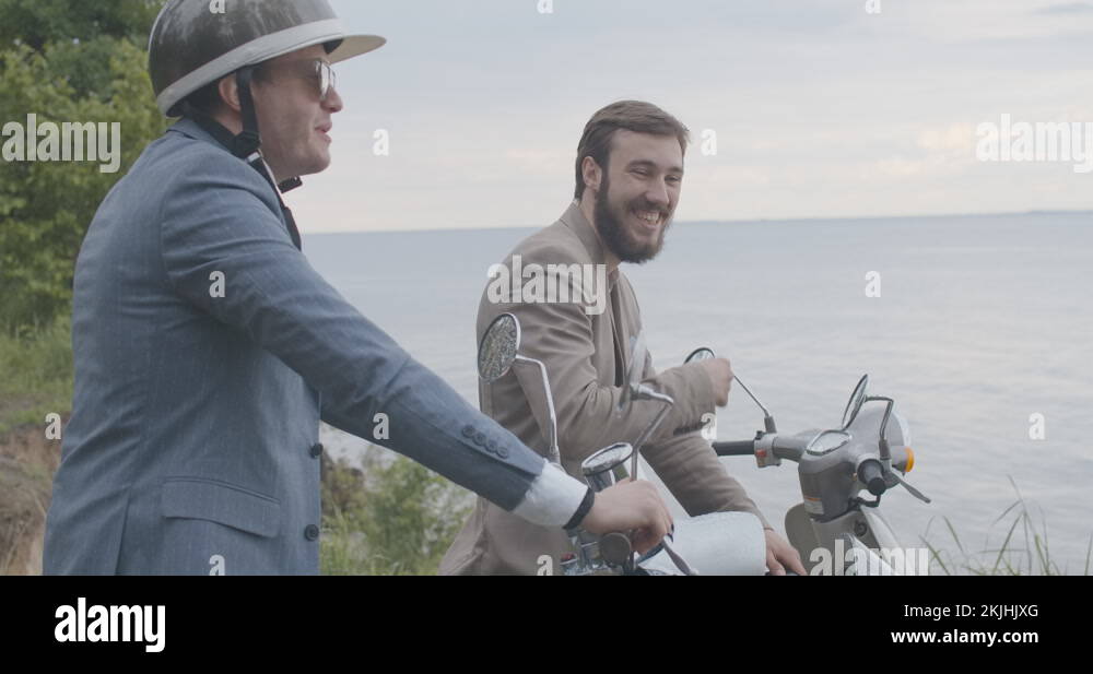 Positive smiling men sitting on scooters on river bank and talking ...