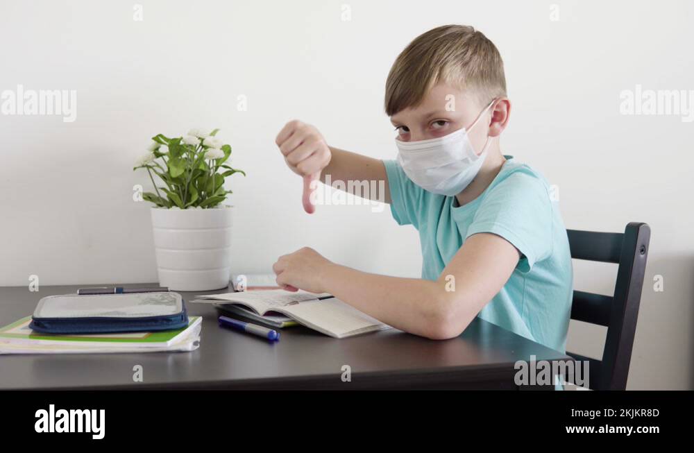 A young boy in a face mask shows a thumb down to the camera and shakes ...
