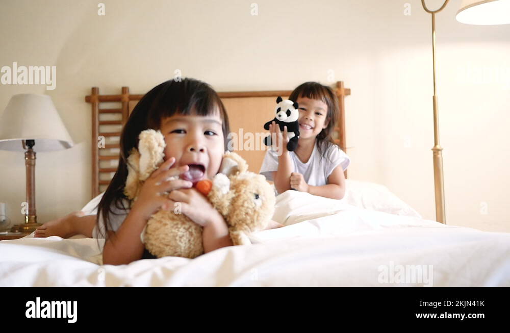 Two little sibling girls in white pajamas are hugging doll lying on bed ...