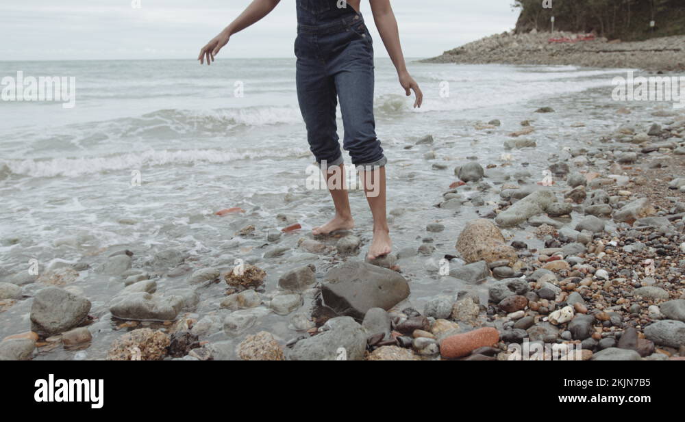 Close up of barefoot female feet in sea stepping on stones at beach ...