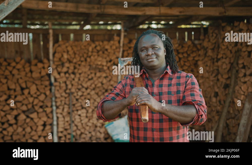 African man lumberjack with the axe standing in front of stack of ...