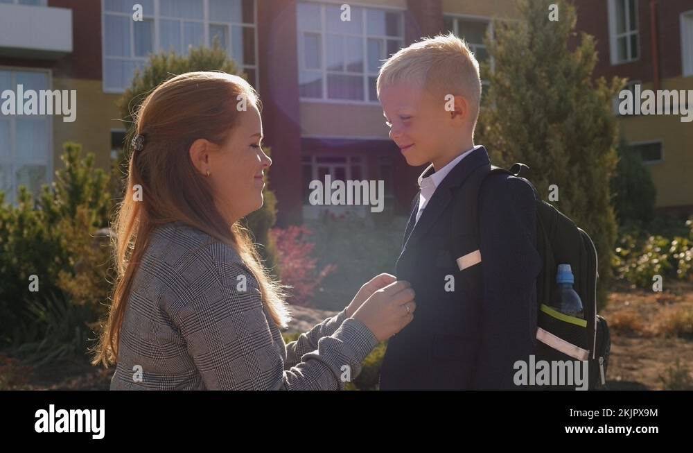 A mother helps her son put on a backpack and buttons his jacket near ...