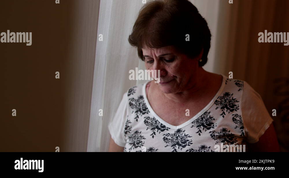 Sad older woman standing by window at home. Depressed worried 60 year ...