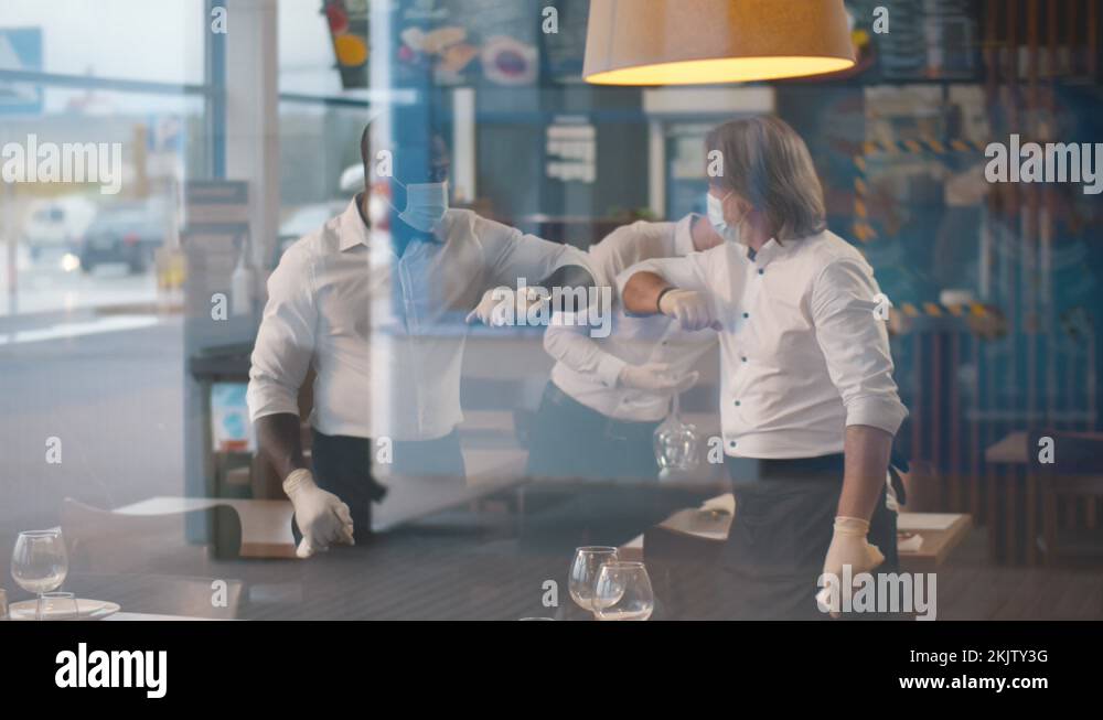 Diverse waiters in medical mask and gloves bumping elbows while setting ...