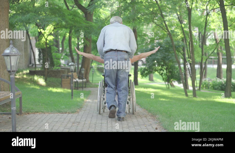 Back view of old man running with wheelchair along the alley as happy ...
