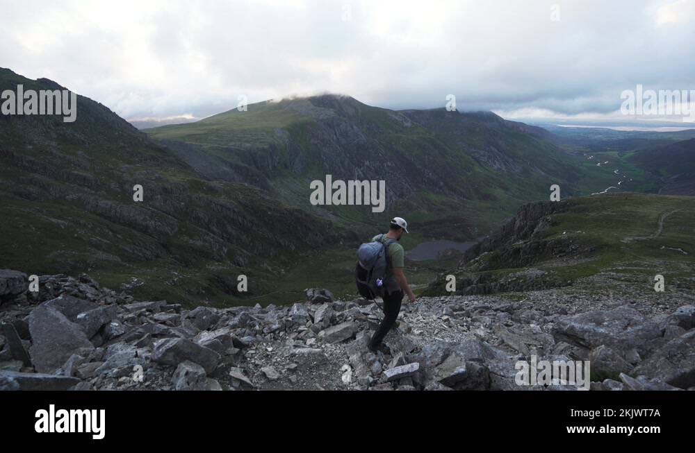A man walking down a steep rocky mountain slope overlooking Llyn Ogwen ...