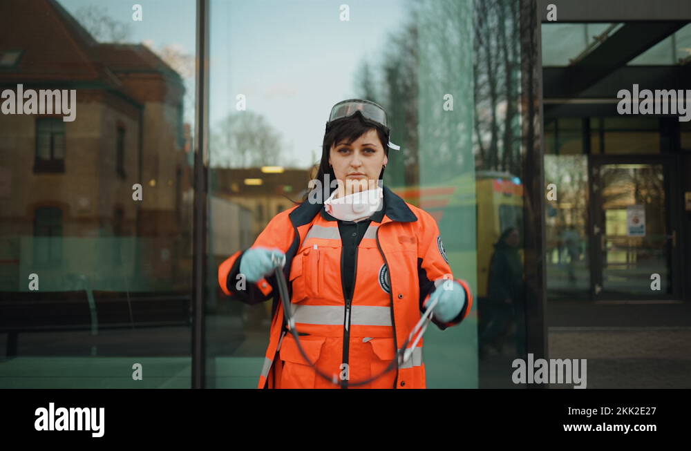 Pretty female paramedic stands near modern hospital and looks at camera ...