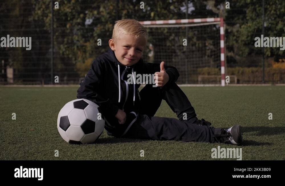 A little boy player sits on a soccer field with a ball and gives a ...